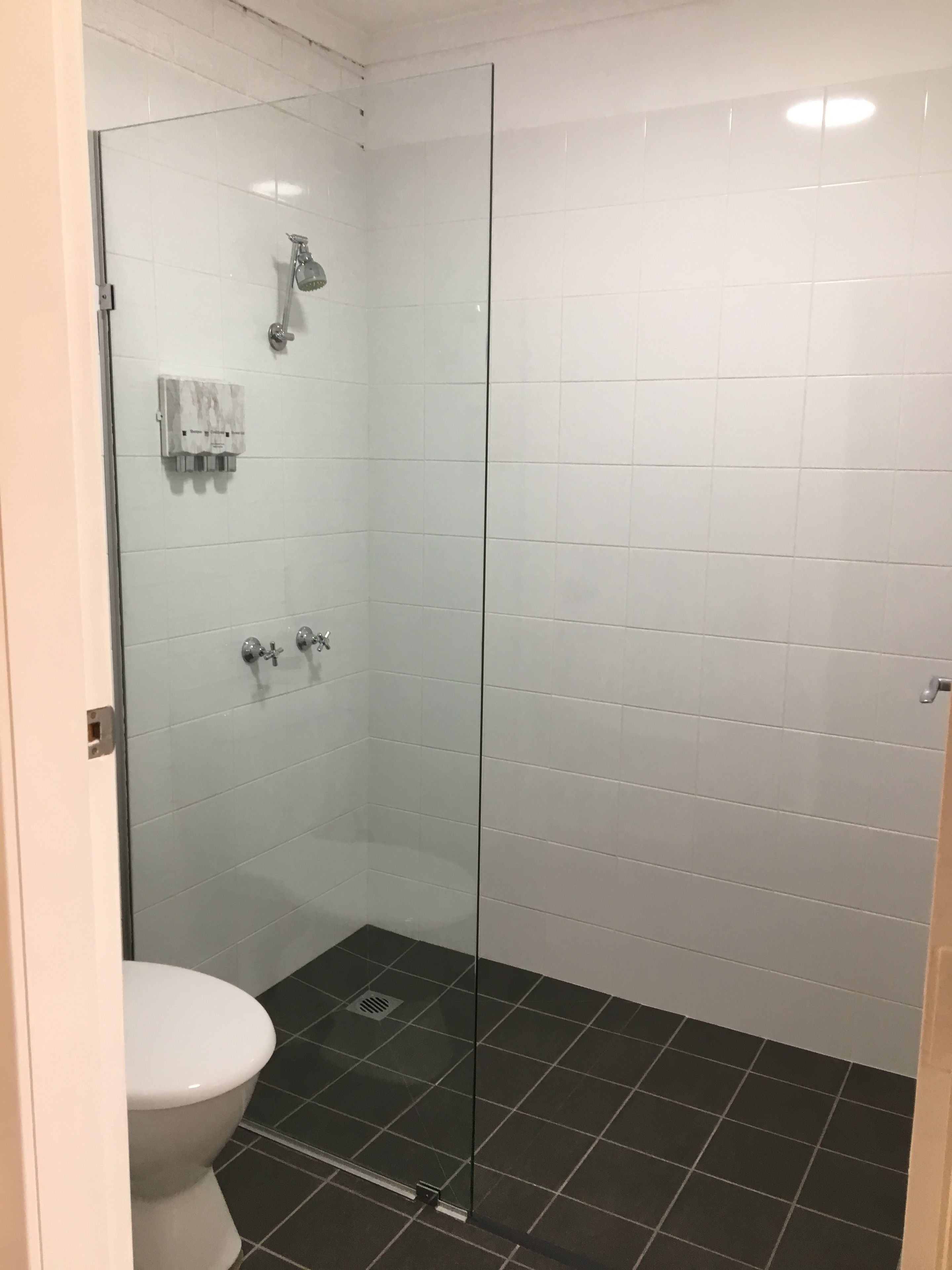 Divisória de chuveiro envidraçada com azulejos brancos, vaso sanitário e dispensador de toalhas.