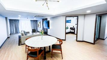 Quarto de hotel moderno com uma mesa de jantar redonda, um sofá, uma TV e uma janela grande com cortinas.