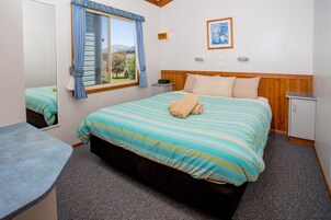 Um quarto com cama, mesa de cabeceira, espelho, janela com cortinas e um pequeno armário.