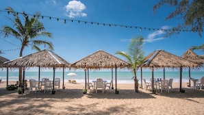 Uma praia com guarda-sóis de palha, cadeiras de plástico brancas e plantas em vasos.