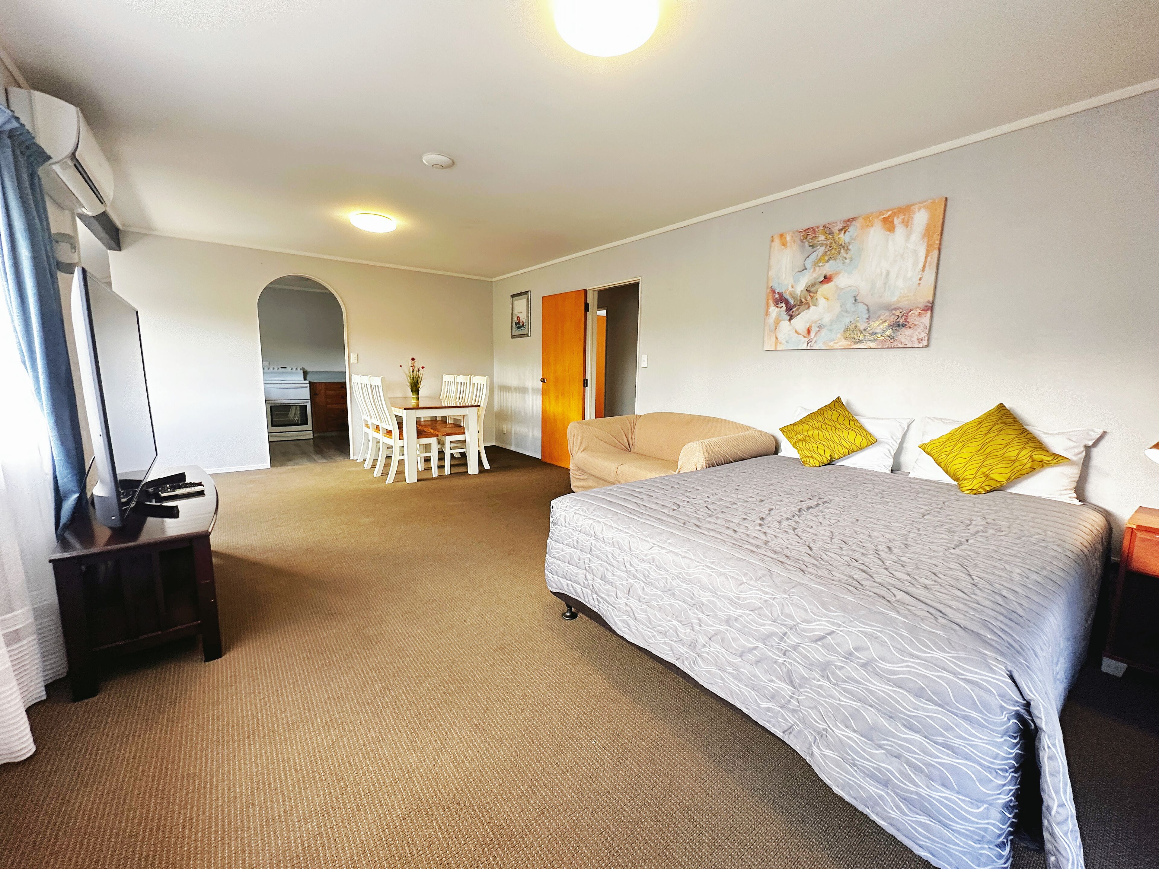 Quarto de hotel com uma cama grande, uma escrivaninha com computador, uma mesa de jantar com cadeiras e uma área de cozinha ao fundo.