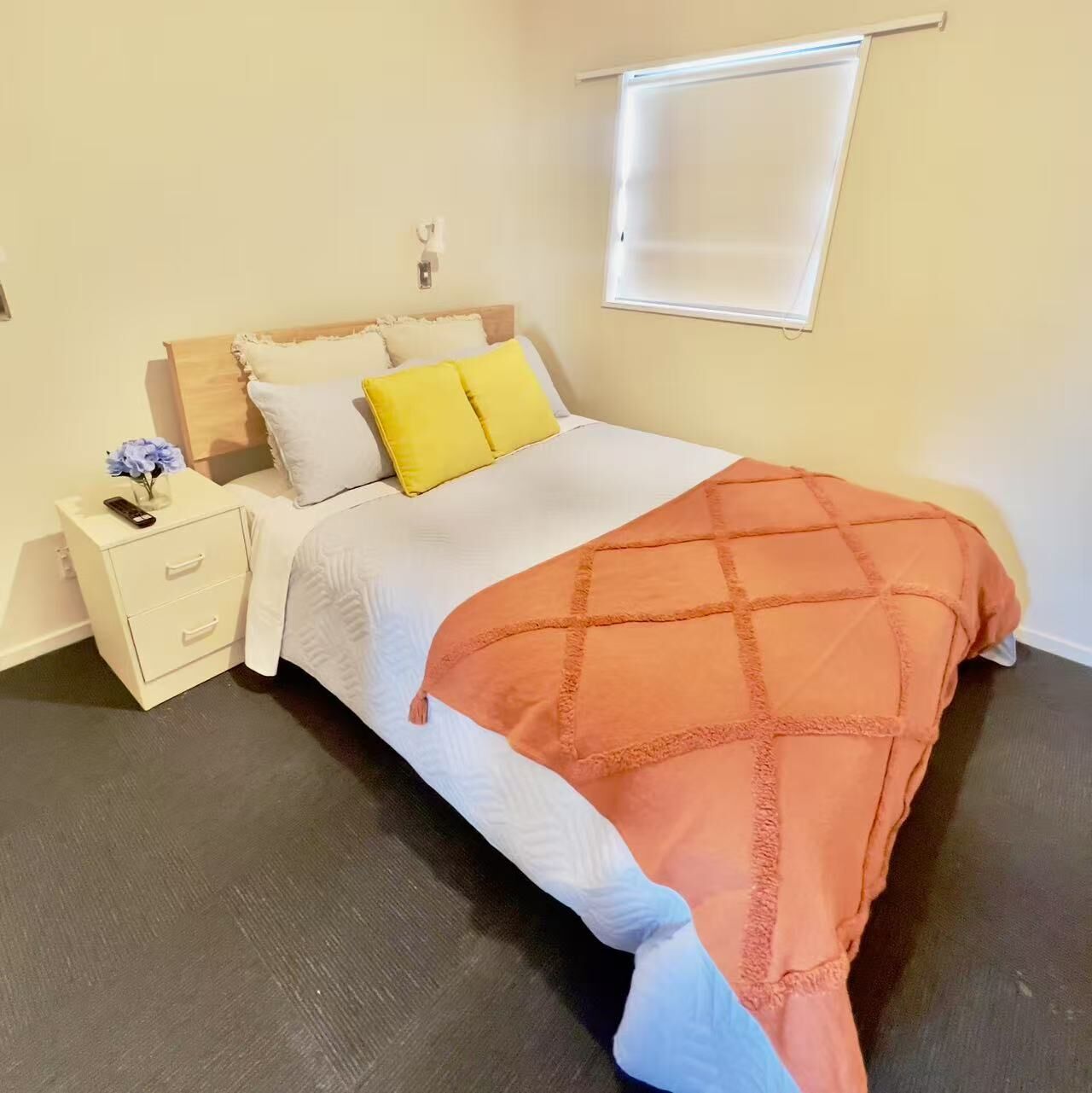 Uma cama bem arrumada com um edredom branco e laranja, uma mesa de cabeceira com abajur e uma janela com persianas.