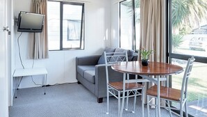 Quarto moderno com sofá cinzento, mesa redonda de madeira com cadeiras, televisão de ecrã plano na parede e amplas janelas com cortinas.