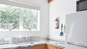 Uma cozinha com armários de madeira, uma geladeira branca, uma janela com vista para o verde e um extintor de incêndio fixado na parede.