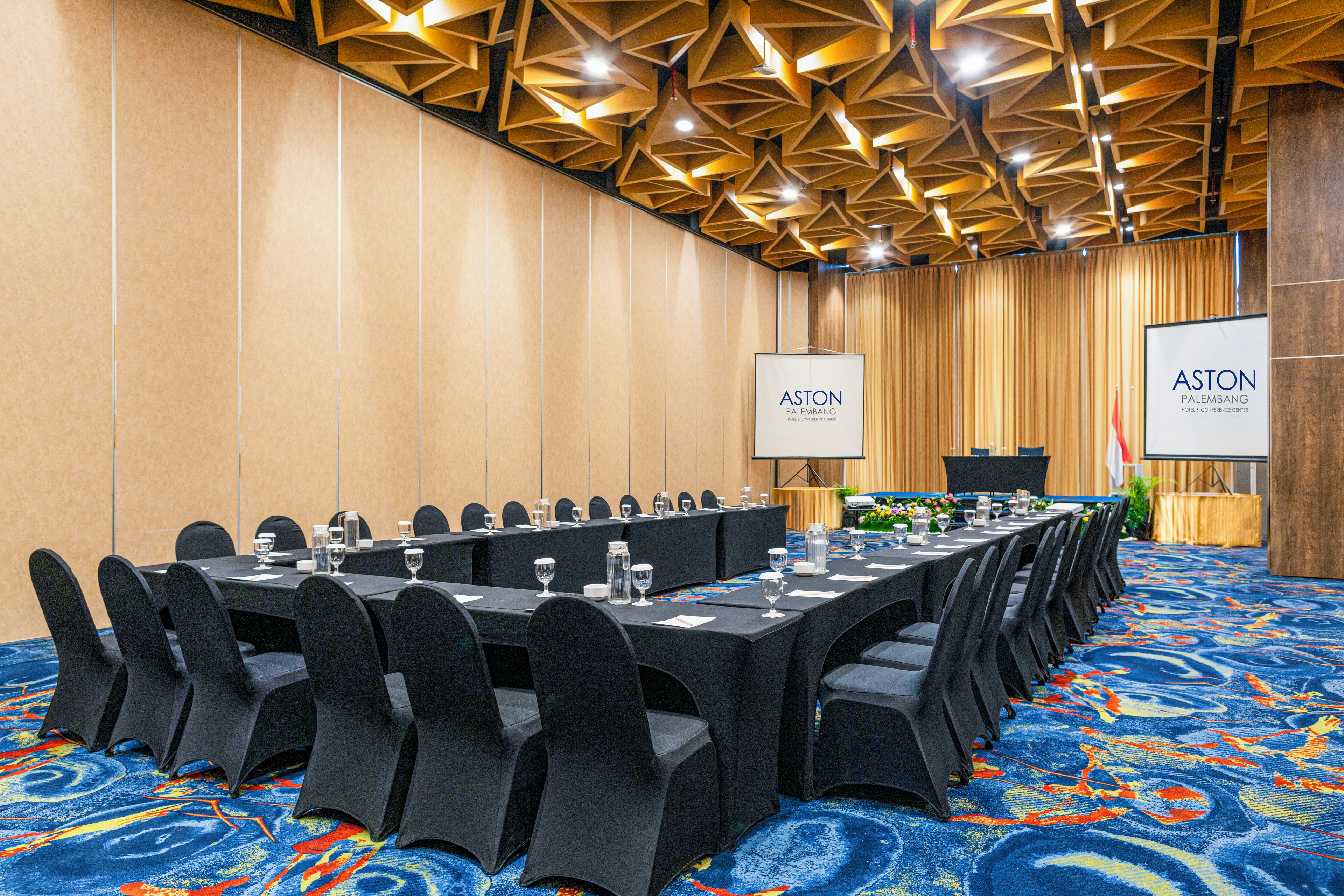 Uma sala de conferências com uma mesa comprida, cadeiras pretas e um tapete azul estampado. Duas telas exibem o logotipo "ASTON PALEMBANG HOTEL & CONFERENCE CENTER".