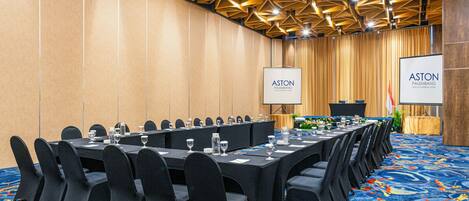 Uma sala de conferências com uma mesa comprida, cadeiras pretas e um tapete azul estampado. Duas telas exibem o logotipo "ASTON PALEMBANG HOTEL & CONFERENCE CENTER".