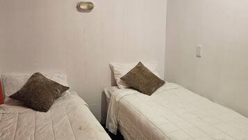 Quarto pequeno e limpo com duas camas individuais, cada uma com uma almofada e mesa de cabeceira.