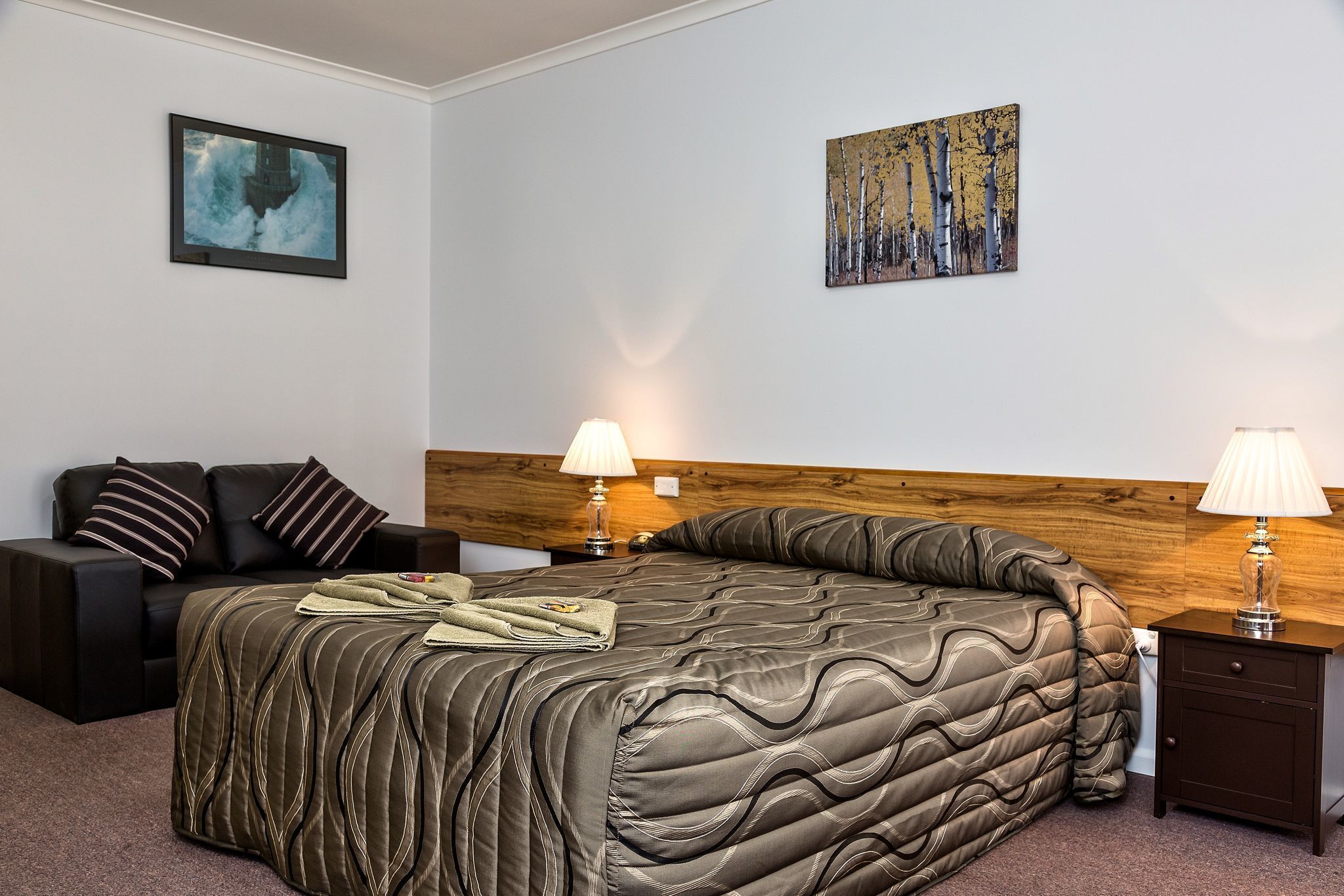 Quarto de hotel com uma cama grande, cabeceira de madeira, duas luminárias de mesa, um sofá preto e duas obras de arte na parede.