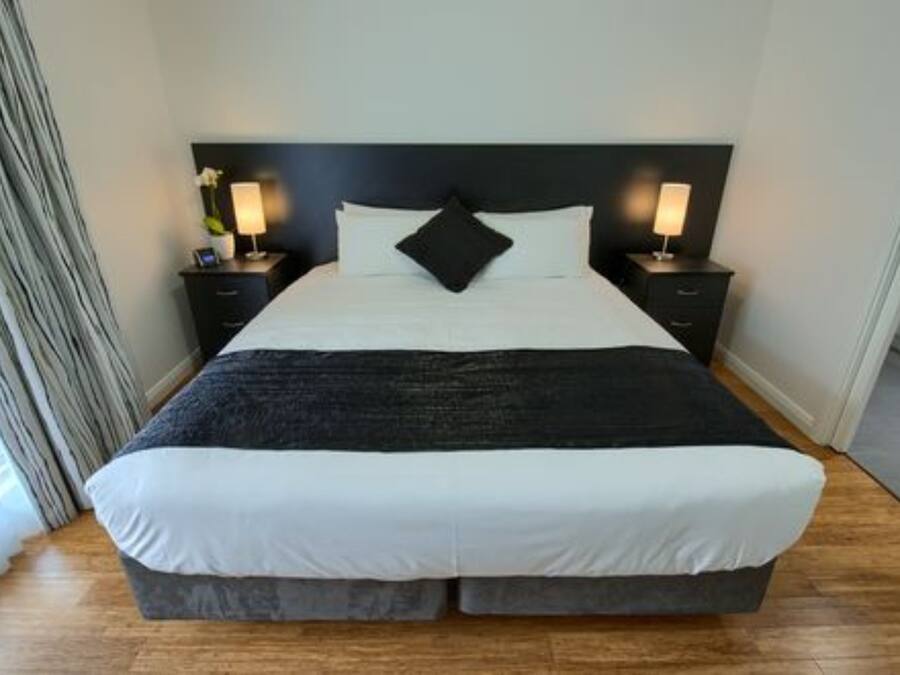 Cama bem arrumada com roupa de cama branca e preta, duas mesas de cabeceira com abajures e uma cabeceira escura.