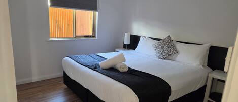 Uma cama bem arrumada com esquema de cores preto e branco, uma mesa de cabeceira com abajur e uma janela com persianas.
