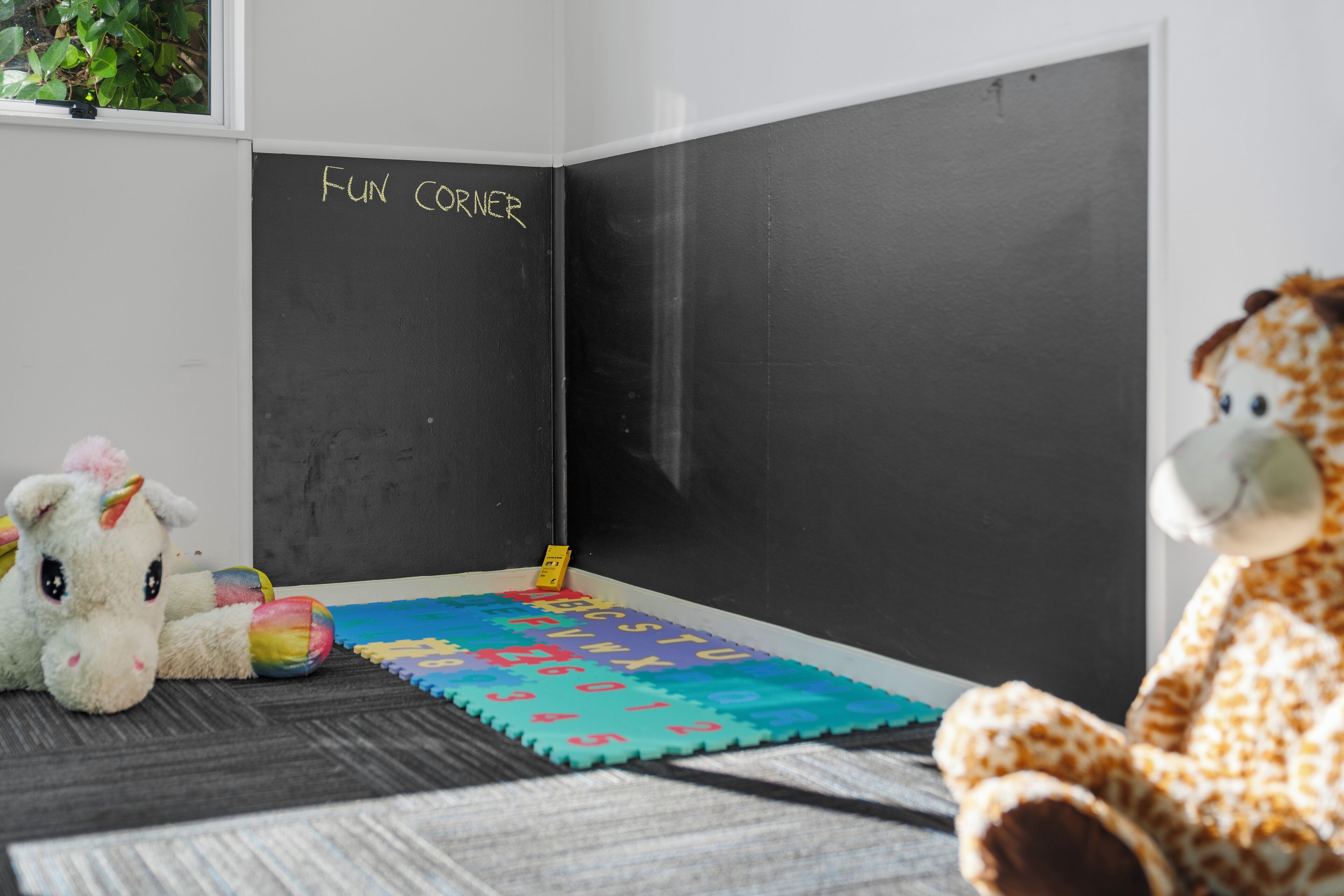 Espaço de recreação infantil com um quadro negro identificado como "FUN CORNER", um brinquedo de girafa de pelúcia e um brinquedo de unicórnio.