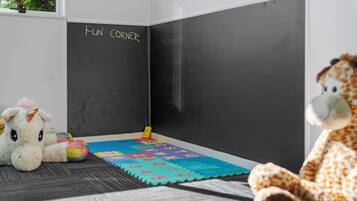 Espaço de recreação infantil com um quadro negro identificado como "FUN CORNER", um brinquedo de girafa de pelúcia e um brinquedo de unicórnio.