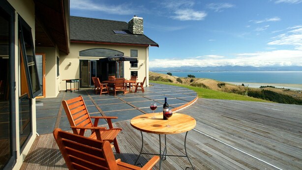 Uma casa moderna com um terraço com vista para a praia, com móveis de exterior e uma mesa com copos de vinho.