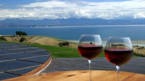 Duas taças de vinho sobre uma mesa de madeira com vista panorâmica para um corpo d'água e montanhas ao fundo.