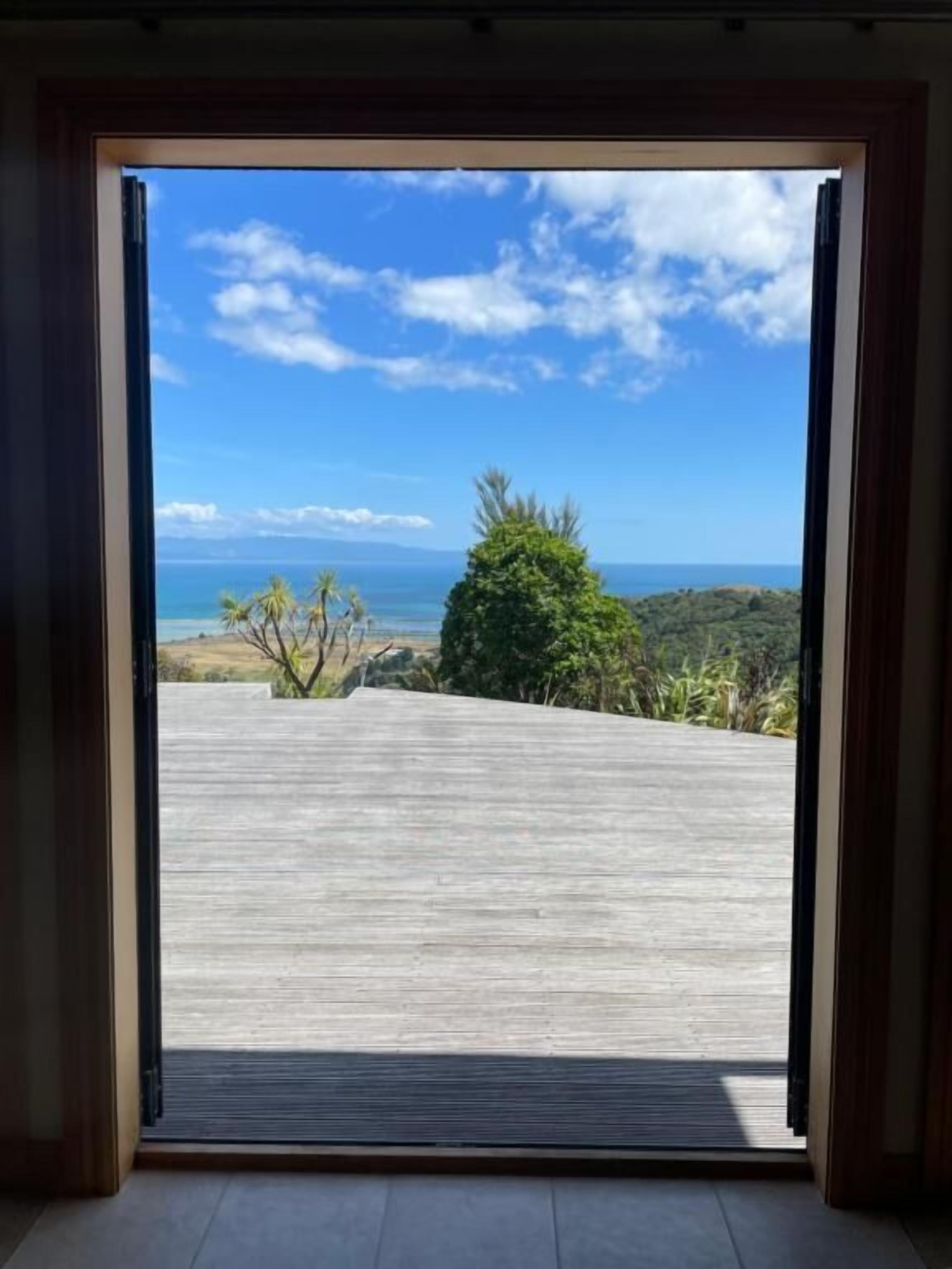 Visão através de uma porta de correr para um deck de madeira, céu claro e paisagem distante.
