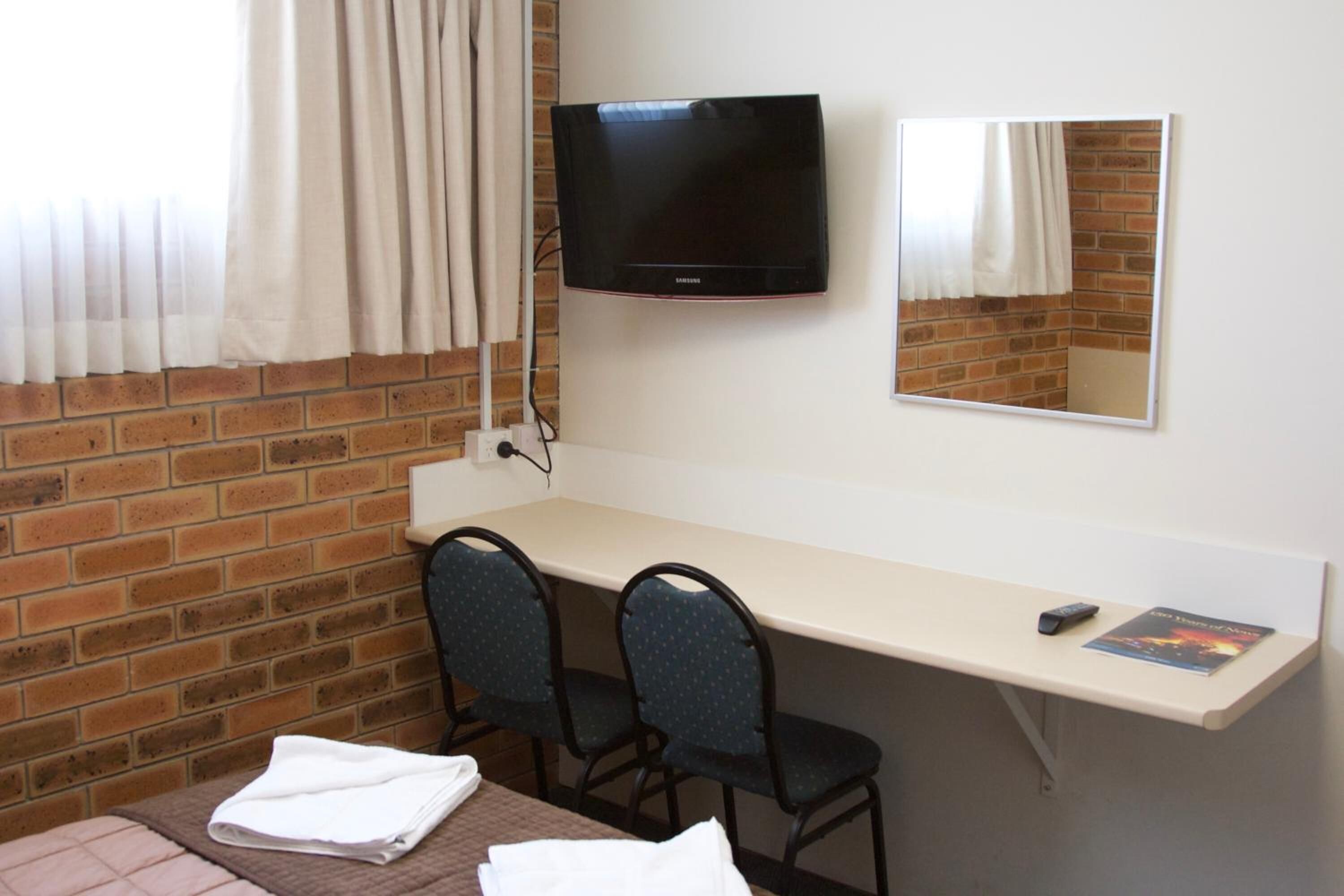 Quarto de hotel com parede de tijolos, televisão de tela plana presa acima de uma escrivaninha, duas cadeiras, uma cama com toalha branca dobrada e um controle remoto na escrivaninha.