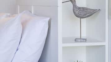 Quarto com uma cama branca, uma mesa de cabeceira branca, uma escultura decorativa de pássaro, uma pequena planta em vaso e uma revista sobre a mesa de cabeceira.