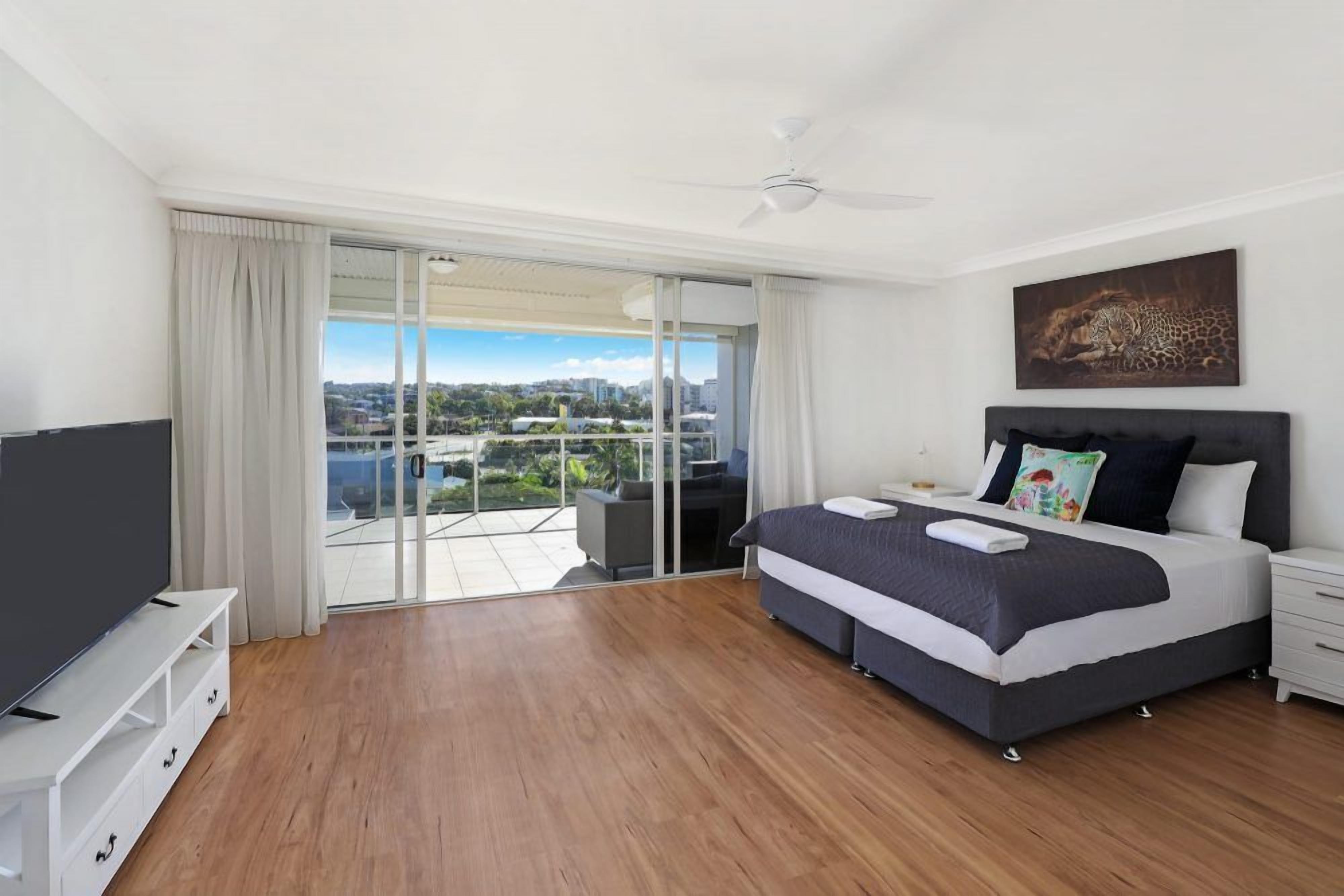 Quarto moderno com uma cama grande, televisão de tela plana, ventilador de teto e vista para a cidade por uma porta de vidro deslizante.