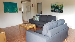 Uma sala de estar com um sofá cinzento, uma mesa de centro de madeira e piso revestido de azulejos.