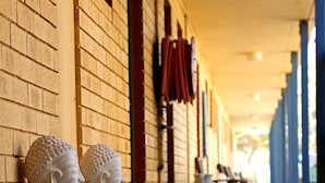 Um corredor com duas estátuas de Buda sentadas em frente a uma parede de tijolos, que leva a uma área coberta com mesas e cadeiras.