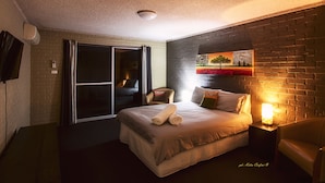 Quarto de hotel com cama, cadeira, mesa de cabeceira com abajur, quadro na parede e porta deslizante que dá para outro quarto.