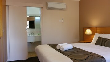 Quarto de hotel com cama, mesa de cabeceira, luminária, ar-condicionado e banheiro visível por uma porta aberta.