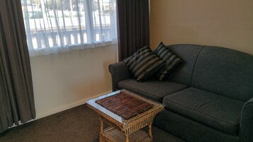 Sala de estar com sofá cinza, duas almofadas listradas, mesa de centro trançada e janela com cortinas leves.