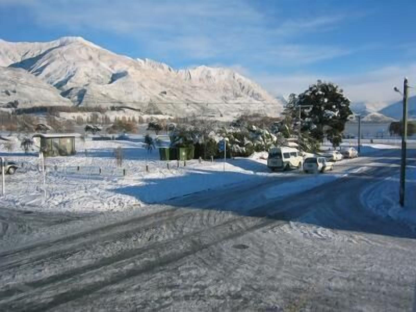 Paisagem nevada com uma montanha ao fundo, alguns carros estacionados e um pequeno edifício.