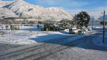 Paisagem nevada com uma montanha ao fundo, alguns carros estacionados e um pequeno edifício.