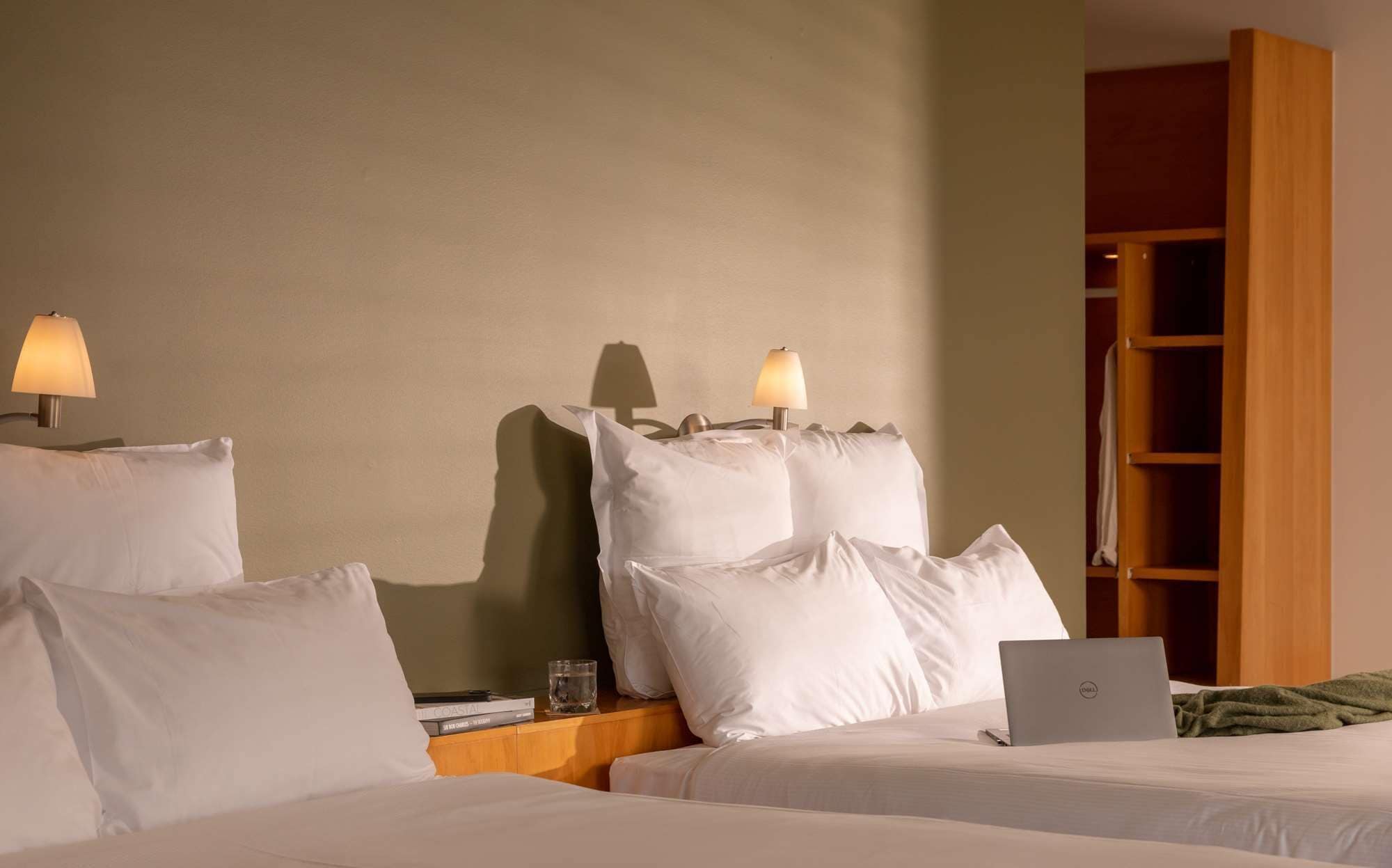 Quarto de hotel com duas camas, um laptop em uma delas, uma mesa de cabeceira com abajur e uma estante de livros.