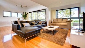 Uma sala de estar moderna com um sofá de couro preto, um sofá bege e uma mesa de centro. Há janelas amplas que proporcionam luz natural e vista para o verde lá fora.