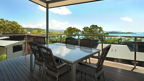 Uma varanda com mesa e cadeiras de vidro, com vista para uma paisagem pitoresca com árvores e um corpo d'água.