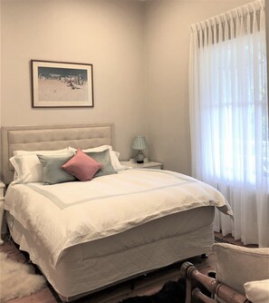 Quarto com cama, mesa de cabeceira, abajur, cadeira e janela com cortinas.