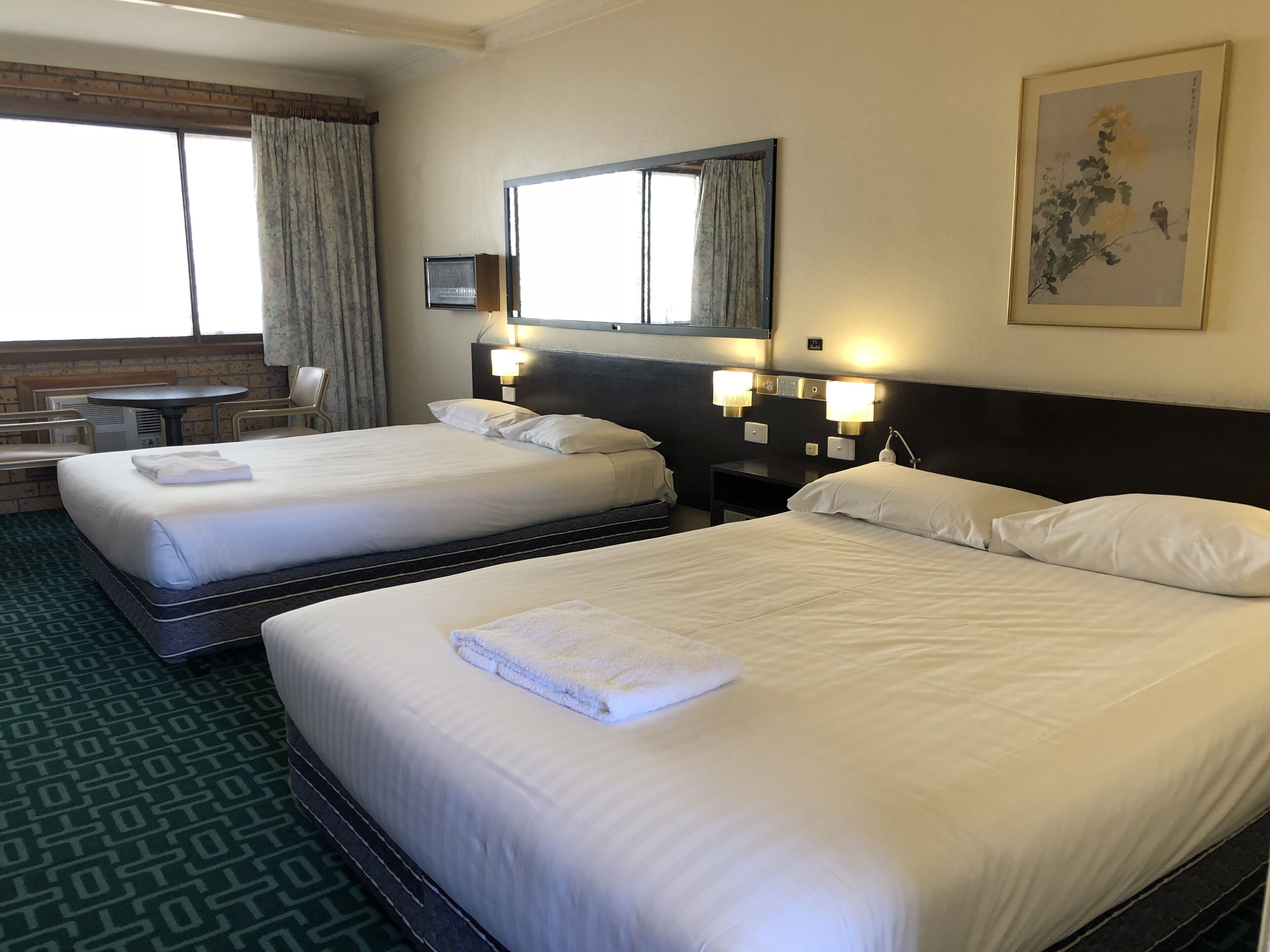 Quarto de hotel com duas camas, uma mesa pequena, uma cadeira, televisão e um quadro na parede.