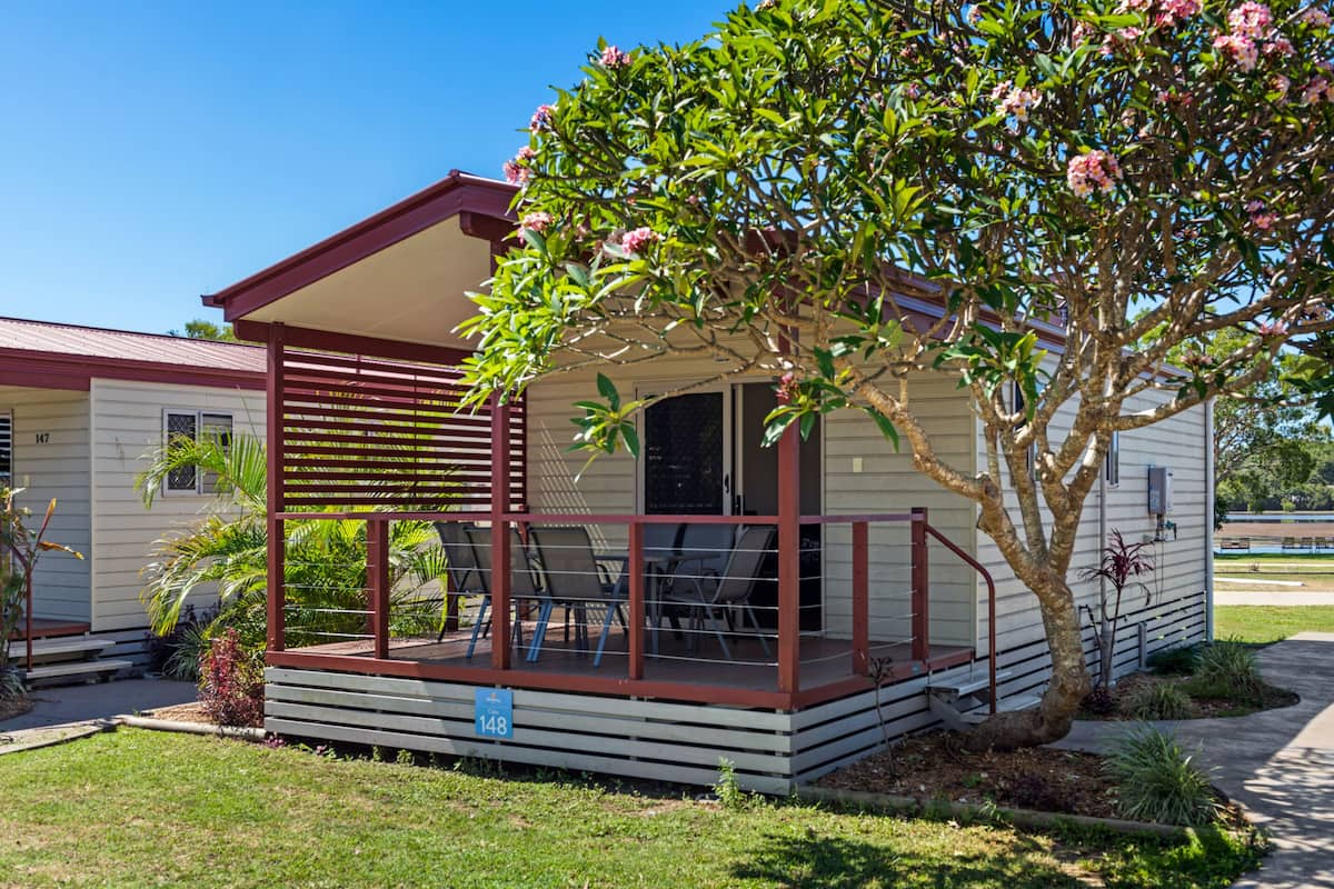 Uma casa pequena e térrea com varanda, uma árvore com flores cor-de-rosa e uma placa com o número 148 no chão.