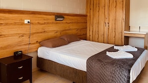 Cama bem arrumada com colcha marrom, mesa de cabeceira com relógio e cabeceira de madeira.