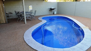 Piscina com borda curva, rodeada por piso de pedras e um espaço coberto com espreguiçadeiras e uma mesa de metal.