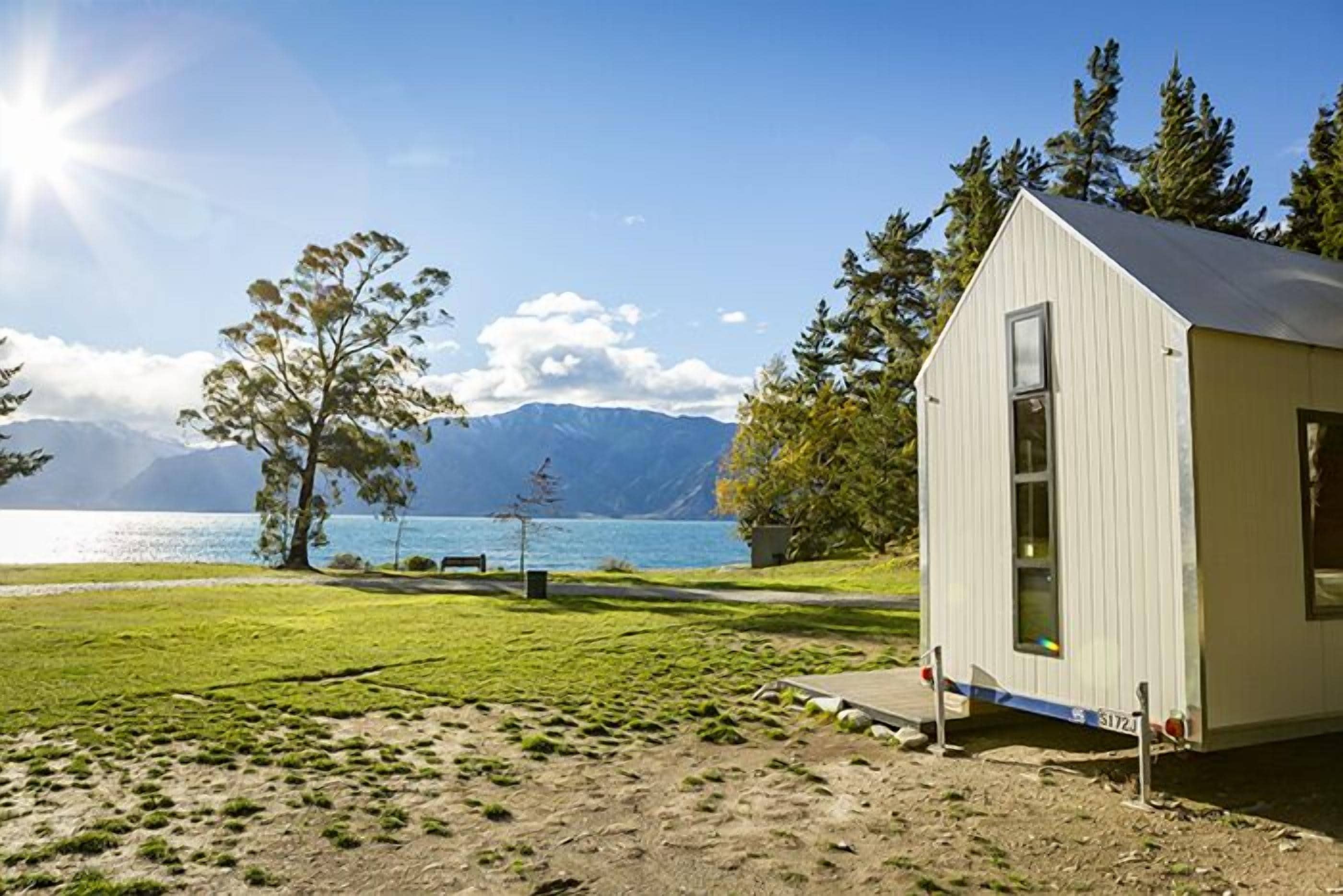 Um pequeno edifício térreo com fachada branca e telhado inclinado, localizado em uma área gramada com vista para um lago e montanhas ao fundo.