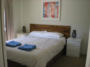Cama bem arrumada com duas toalhas dobradas, cabeceira de madeira, mesa de cabeceira com abajur e uma obra de arte na parede.