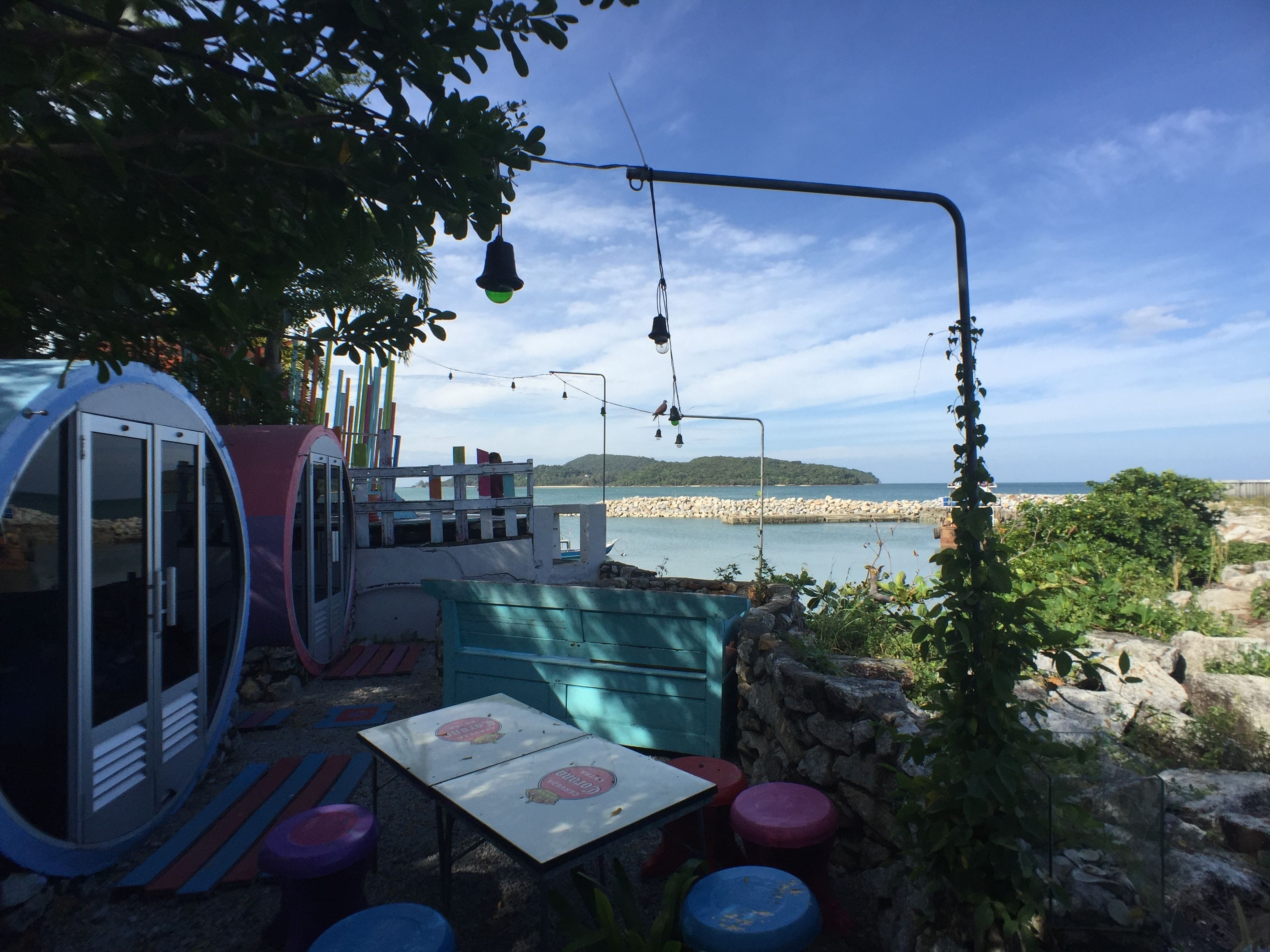 Uma área de estar ao ar livre à beira-mar, com mesa e banquinhos, uma pequena estrutura de recreação e vista para o mar e um litoral rochoso.