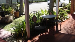 Uma área de estar coberta ao ar livre, com cadeiras e mesas de metal, cercada por plantas em vasos e um deck de madeira.