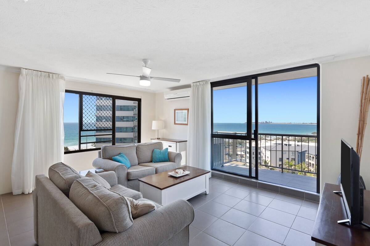 Uma sala de estar moderna com sofá, mesa de centro e televisão. Possui grandes janelas com vista para a praia e oceano.