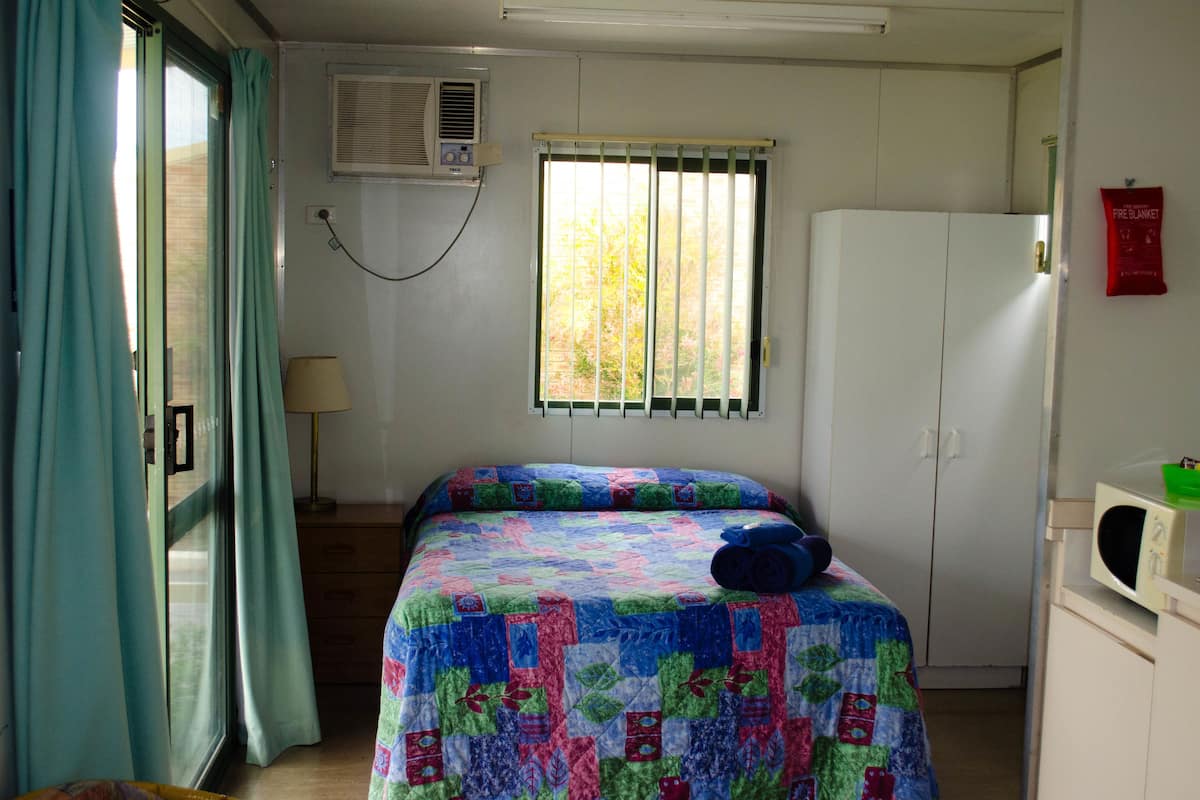 Quarto com cama, janela com persianas, micro-ondas, manta anti-fogo e uma pequena mesa com abajur.
