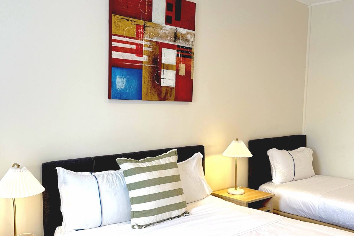 Quarto de hotel com uma cama grande, duas mesinhas de cabeceira, uma luminária e um quadro abstrato colorido na parede.