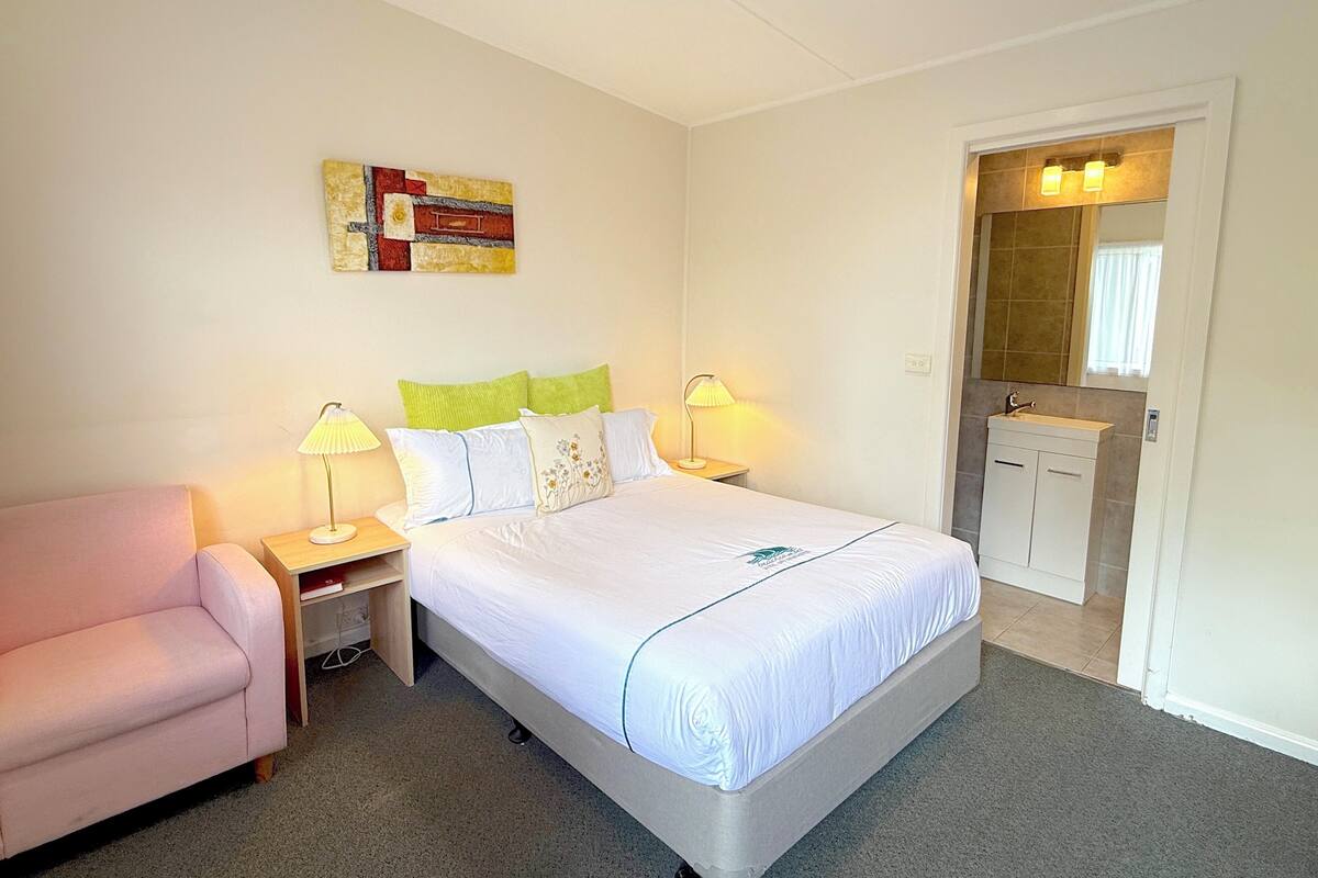 Quarto de hotel com cama, mesas de cabeceira, luminária, cadeira, banheiro com espelho e uma obra de arte na parede.
