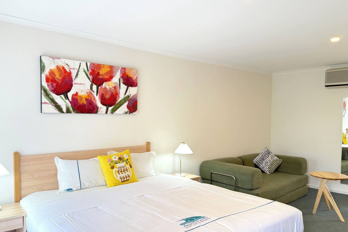 Quarto de hotel com uma cama, um sofá verde, uma pequena mesa de apoio de madeira e uma pintura de tulipas na parede.