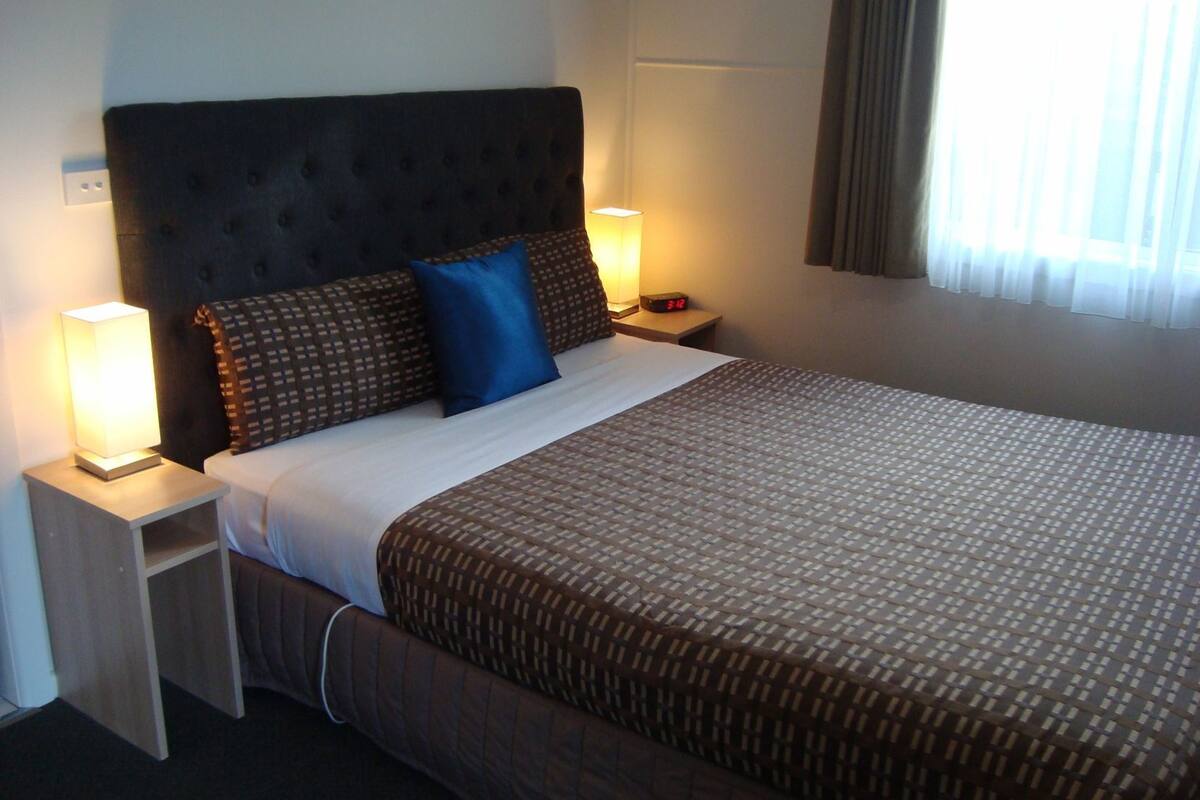 Quarto de hotel com cama grande, duas luminárias de cabeceira, mesa de cabeceira e janela com cortinas.