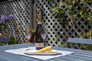 Uma garrafa de vinho Jansz Tasmania, duas taças e uma fatia de queijo sobre uma mesa, com uma cerca treliçada ao fundo.