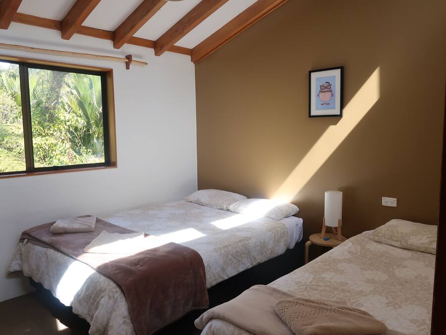 Quarto com duas camas de solteiro, janela com vista para área verde, quadro na parede e mesa de cabeceira com abajur.