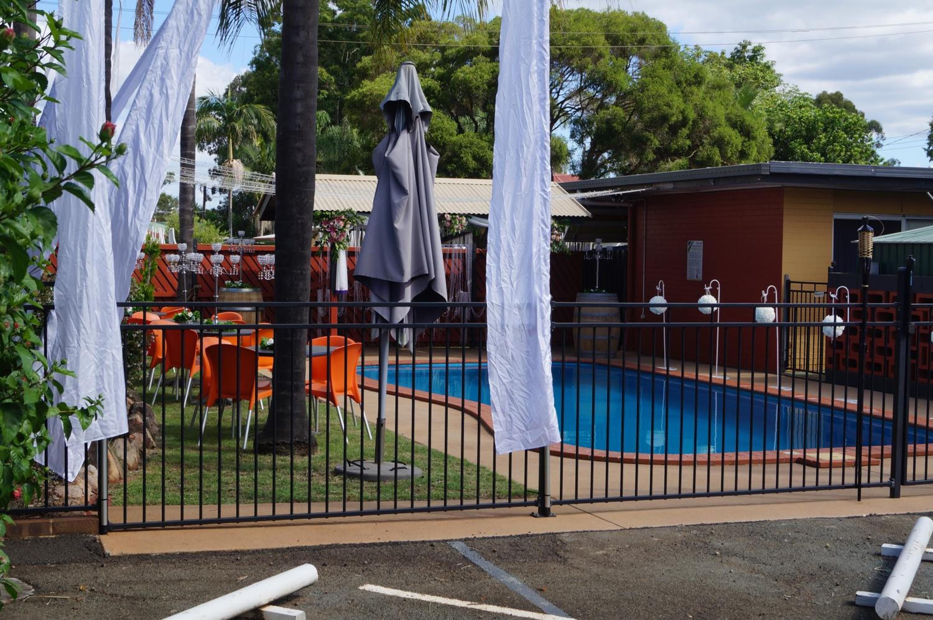 Área de piscina cercada, com cadeiras laranja e uma mesa coberta, rodeada por árvores e um edifício com fachada vermelha e amarela.